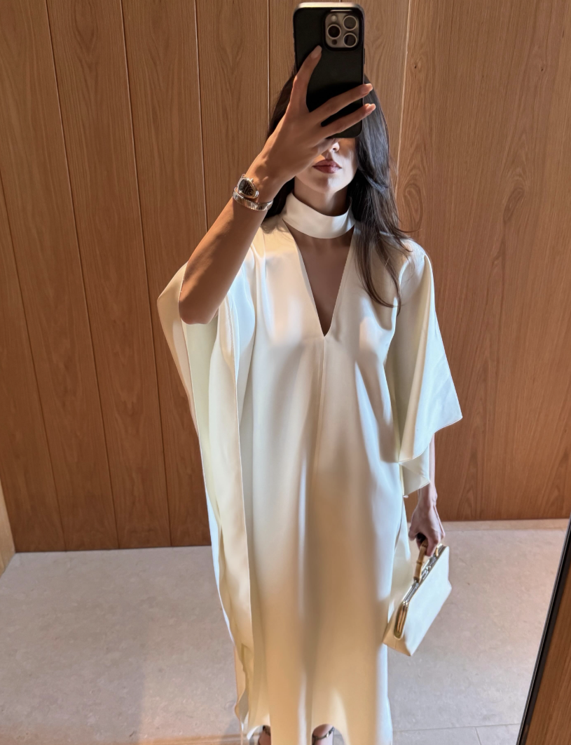 A woman in an elegant white dress takes a mirror selfie. The dress has flowy sleeves and a choker neckline. She holds a small purse%2C conveying a chic%2C sophisticated style.